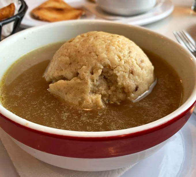 A matzo ball floats serenely in golden broth, looking like edible comfort wrapped in a warm, savory hug.