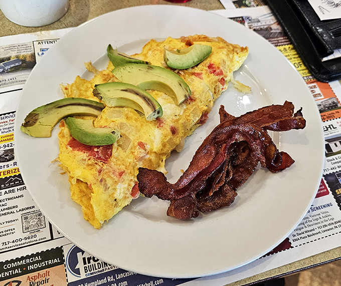 An omelet that's seen the world, with avocado slices lounging on top like they're at a spa retreat. Bacon stands guard nearby.