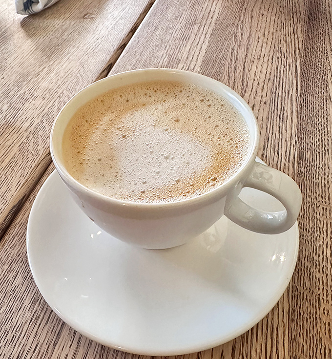 A proper cappuccino served in white porcelain against weathered wood &ndash; the morning ritual that separates "just waking up" from "actually being awake."
