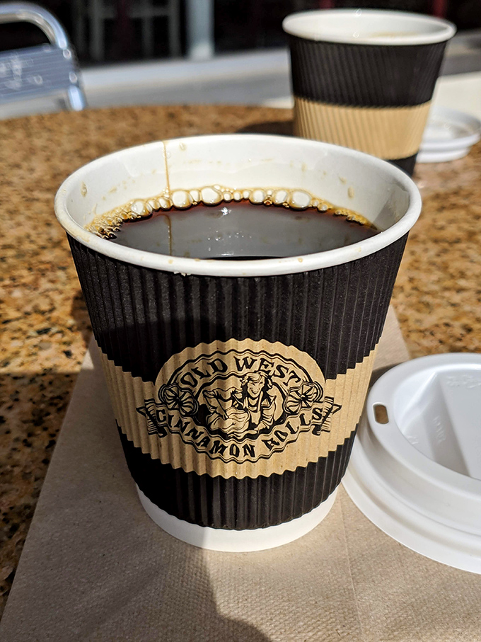 Coffee in its proper habitat&mdash;beside a cinnamon roll. The cup sleeve doubles as a reminder of where to return tomorrow morning.