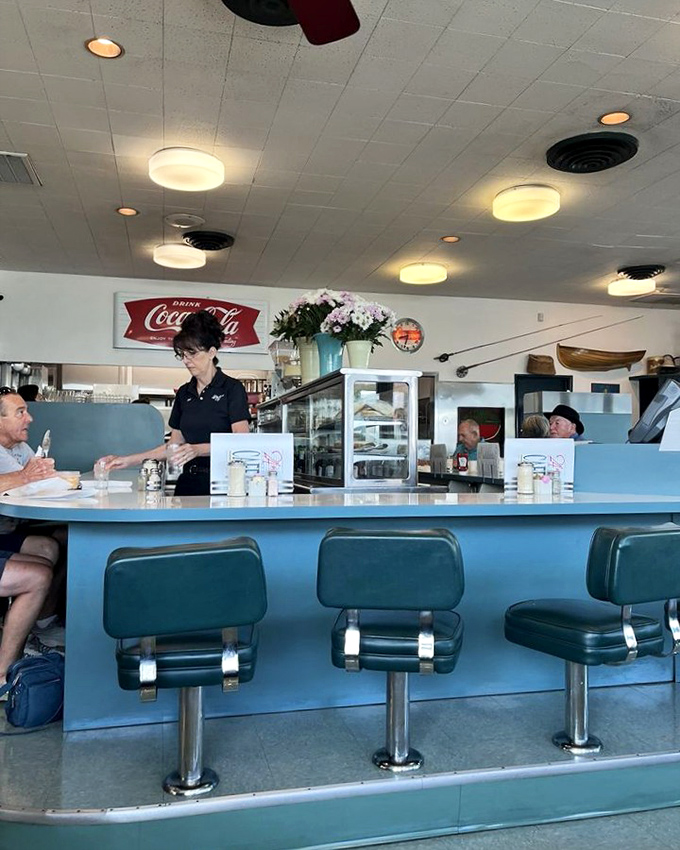 The classic diner counter—where solo diners find community, coffee cups are never empty, and the Coca-Cola sign promises refreshment.