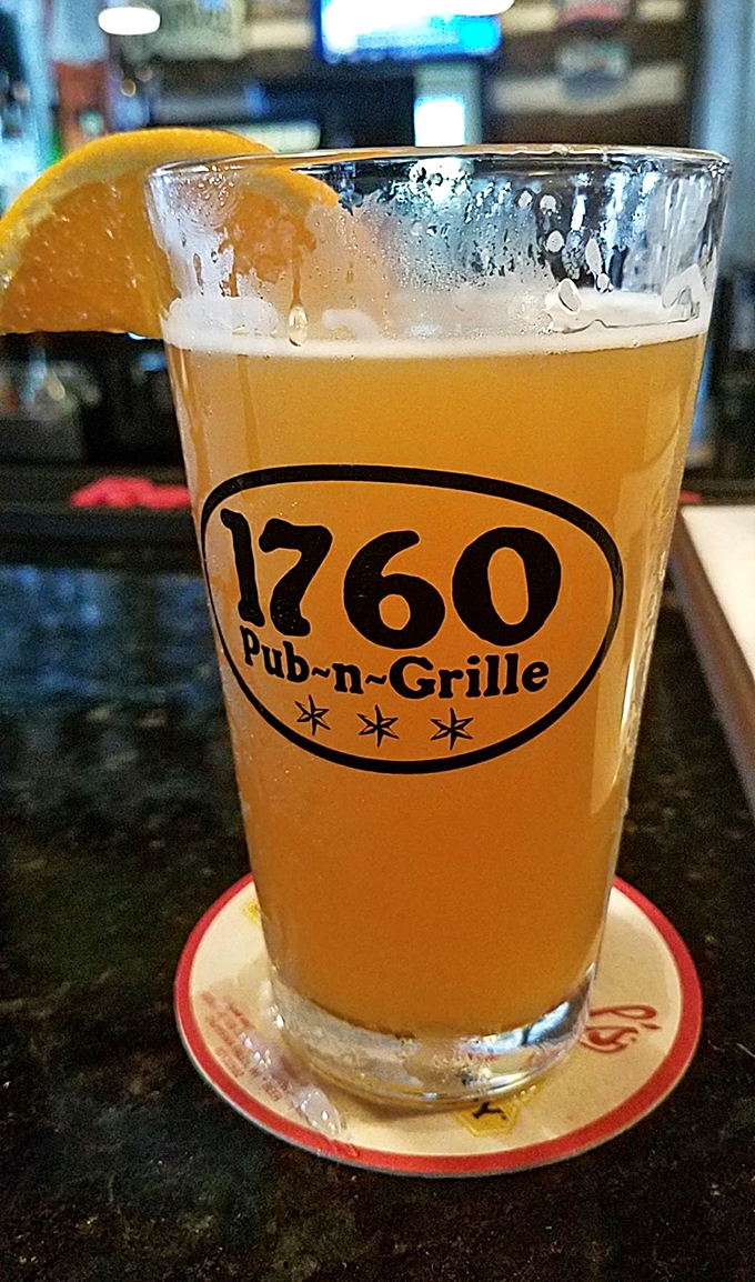 A perfect pour of golden craft beer, adorned with an orange slice, sits in a branded glass &ndash; liquid sunshine waiting to brighten your afternoon.