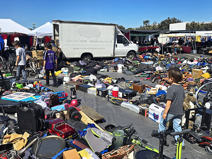 A bustling flea market filled with secondhand treasures&mdash;electronics, toys, tools, and clothes&mdash;sprawled out on tarps as families and bargain hunters browse under a bright blue sky.
