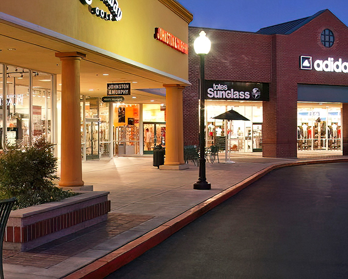 As dusk falls, the outlets transform into a softly lit shopping village where credit cards emerge like nocturnal creatures.