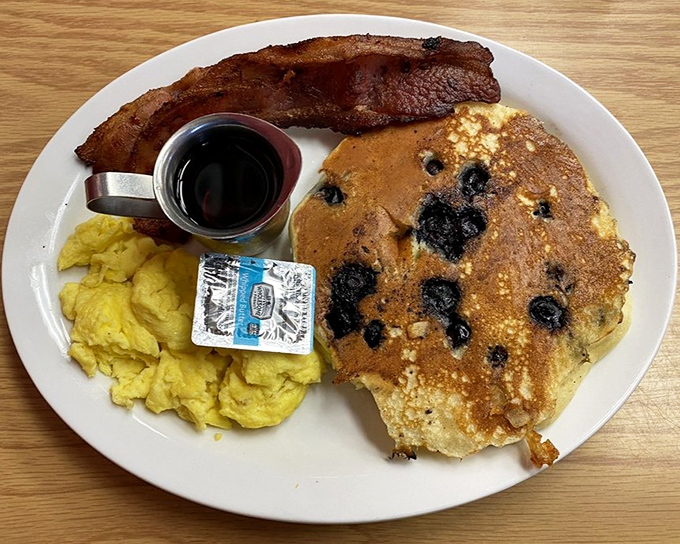 The breakfast trinity: golden blueberry pancake, fluffy scrambled eggs, and bacon cooked to that perfect point between chewy and crisp.