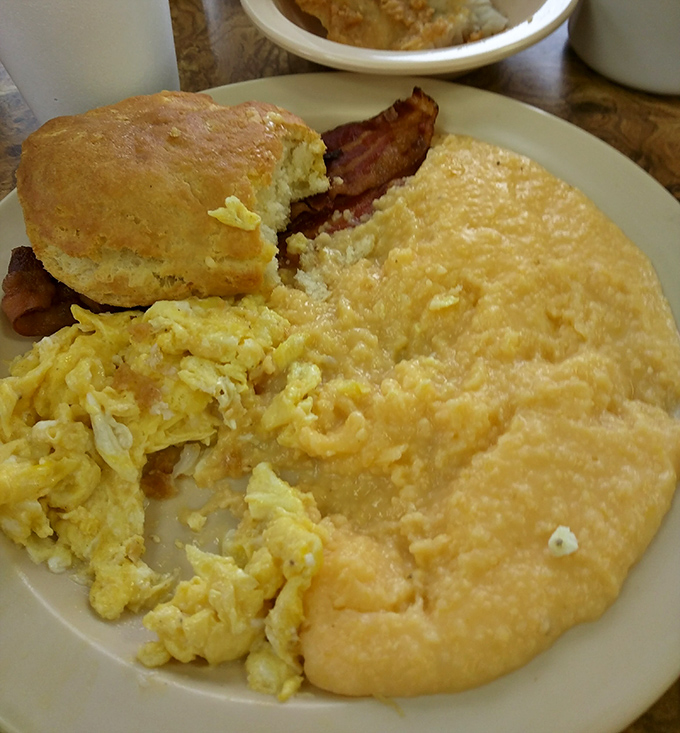 The breakfast trinity: eggs, grits, and bacon with a biscuit standing by &ndash; morning salvation on a simple white plate.