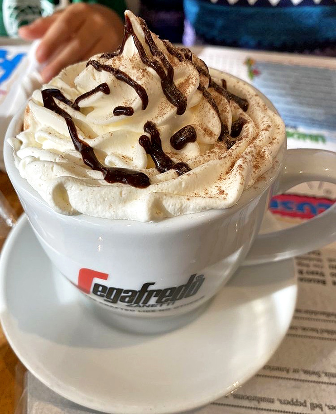 Hot chocolate topped with a cloud of whipped cream and chocolate drizzle – childhood comfort with a grown-up execution.