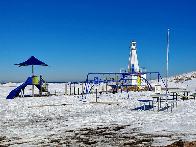 Winter transforms the beach playground into a snow-dusted wonderland where the lighthouse watches over empty swings waiting for summer's return.