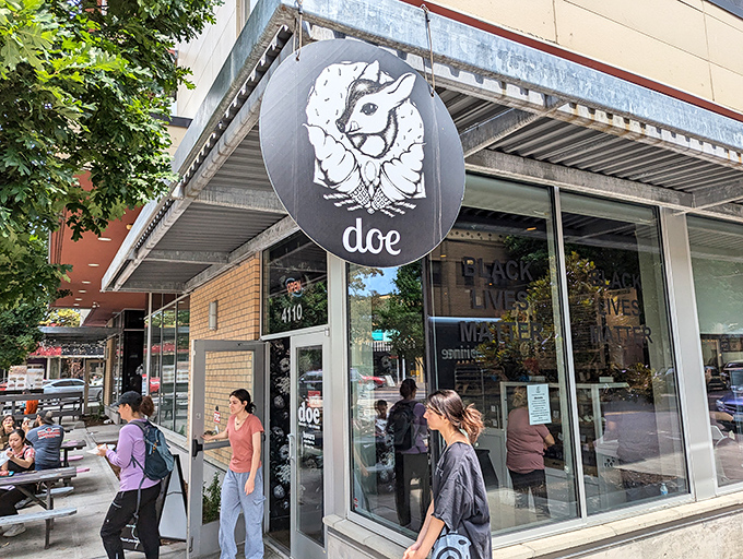Doe Donuts' sleek, modern storefront with its distinctive deer logo brings artisanal donut culture to Portland's Sandy Boulevard. The busy sidewalk shows its popularity.