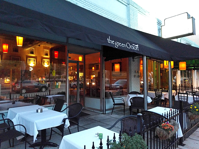 The unassuming storefront of The Green Onion glows with warm invitation at dusk, its patio tables promising culinary adventures within.