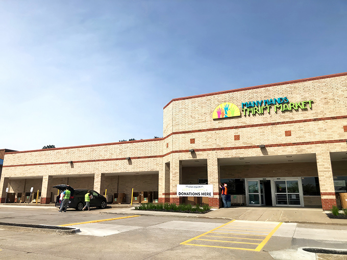 The colorful logo of Many Hands Thrift Market brightens the tan brick exterior, promising treasure hunting adventures within these unassuming walls.