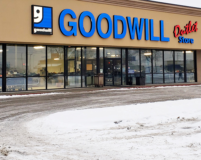 The blue Goodwill Outlet sign beckons like a siren call to treasure hunters. This unassuming exterior houses an indoor archaeological dig waiting to happen.