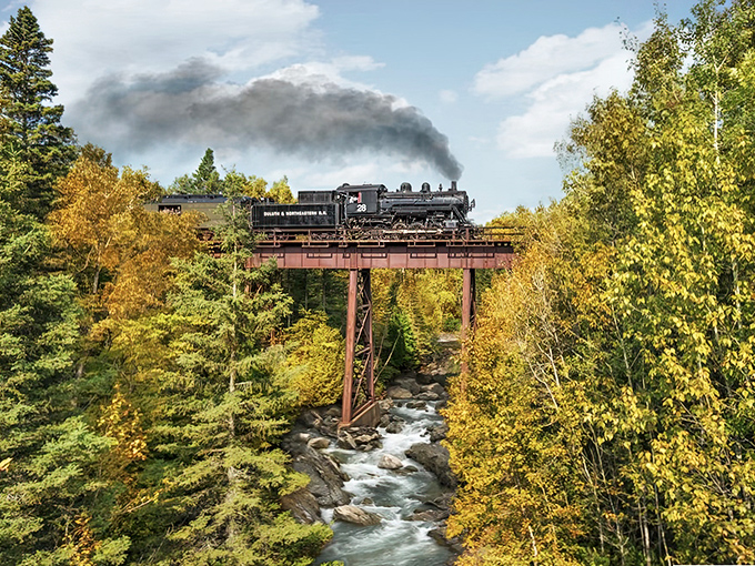The ultimate Minnesota postcard moment: a vintage locomotive crossing a rust-colored trestle bridge over rushing waters, surrounded by autumn's golden canvas.