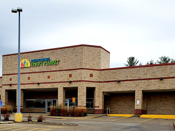 The colorful logo of Many Hands Thrift Market brightens the tan brick exterior, promising treasure hunting adventures within these unassuming walls.