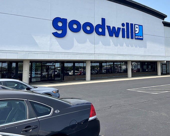 The blue Goodwill sign beckons like a lighthouse for bargain hunters. This unassuming exterior houses a universe of possibilities and second chances.