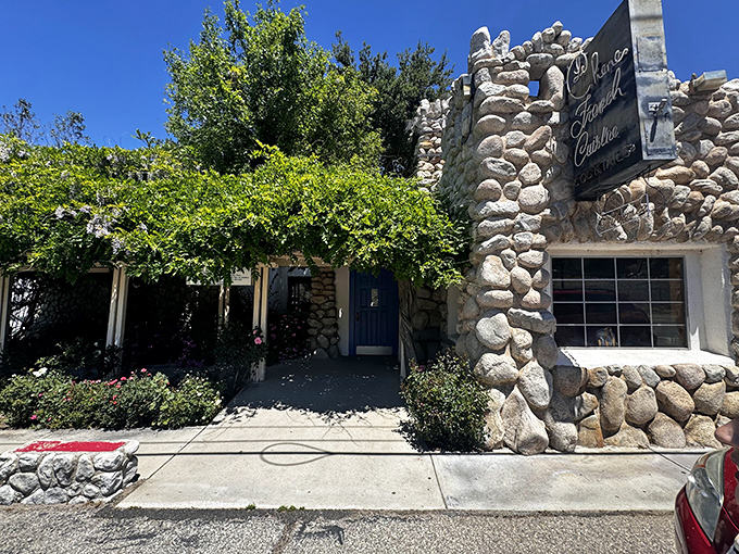 Sunshine bathes the iconic blue door entrance, where climbing vines and blooming flowers offer a warm California welcome with French sensibilities.
