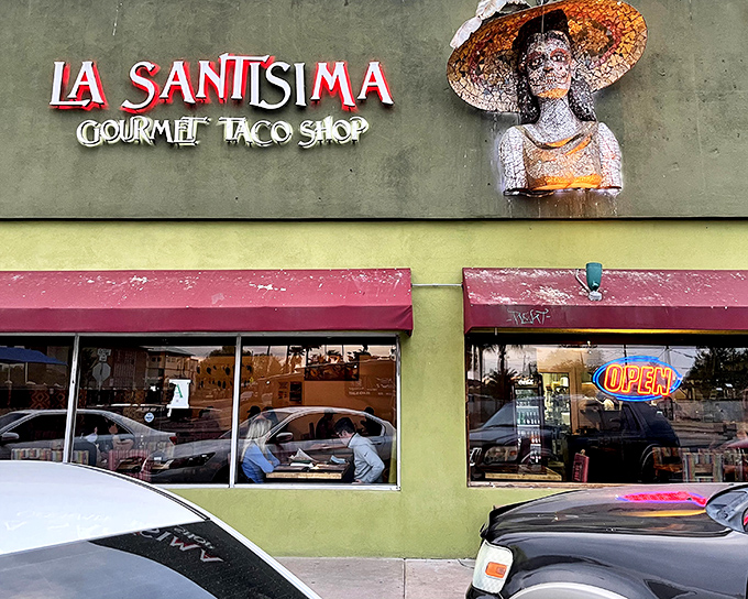 The vibrant exterior of La Santisima beckons like a desert mirage, promising authentic Mexican flavors beneath that distinctive sculptural figure watching over hungry patrons.