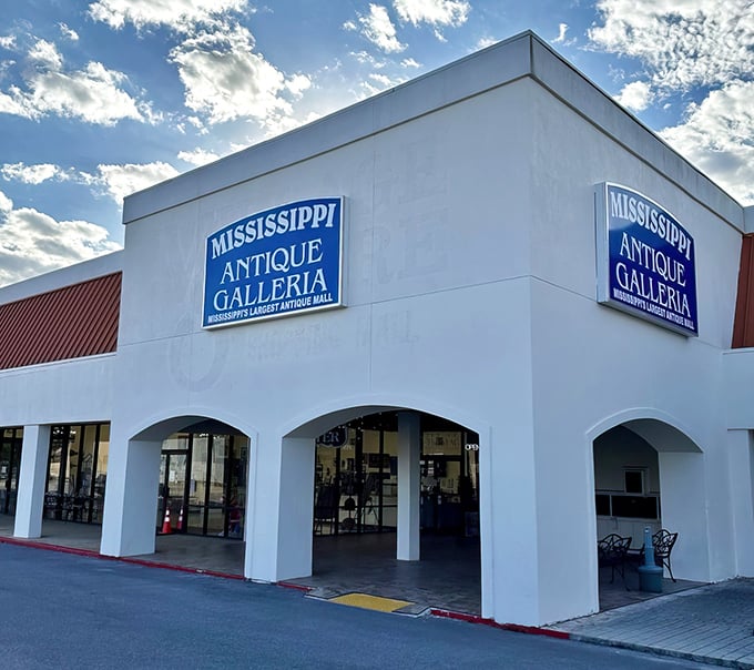 The white facade of Mississippi Antique Galleria stands like a portal to the past, promising treasures behind those blue signs and arched entryways.
