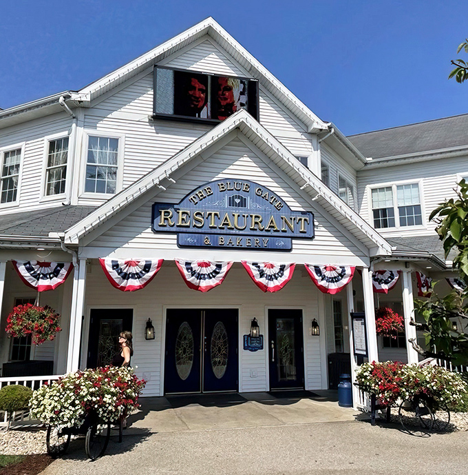 The iconic white facade of Blue Gate Restaurant & Bakery welcomes visitors with patriotic bunting and vibrant flower displays, promising comfort food paradise within.