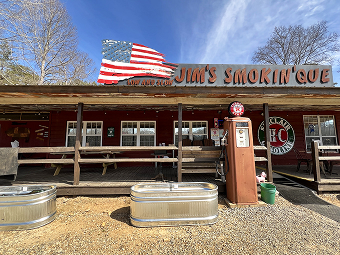 The weathered exterior of Jim's Smokin' Que, complete with vintage gas pump and American flag, promises authentic barbecue without pretension.