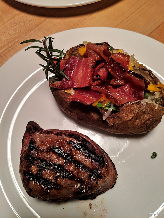A perfectly grilled steak beside a loaded baked potato that's wearing bacon like a fashionable scarf. Comfort food dressed in its Sunday best.