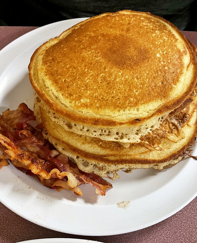 Golden pancakes stacked like edible architecture, accompanied by crispy bacon that knows its job and does it perfectly well.
