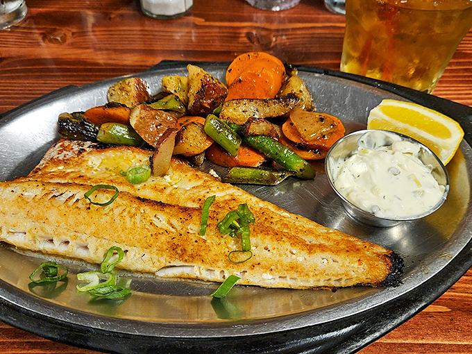 This perfectly seared fish rests on a metal platter like aquatic royalty, surrounded by roasted vegetables that know their supporting role and play it beautifully.