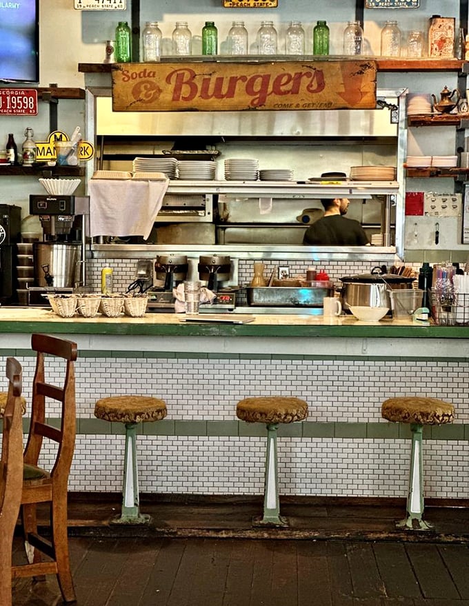 The counter seating offers front-row tickets to the culinary show. Vintage soda bottles and weathered signs create a museum of nostalgic Americana.