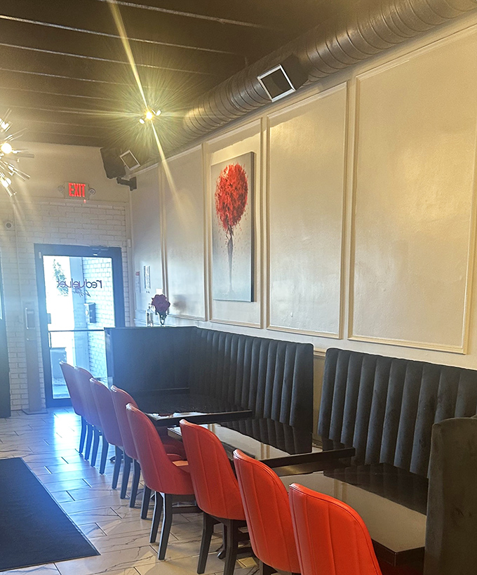 Sunlight streams through the entrance, illuminating a sophisticated space where red accent chairs pop against black booths and marble-look floors.