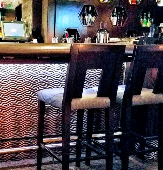 The wave pattern on the bar front catches the light like ripples on a martini. This is where stories begin, end, and occasionally get embellished.