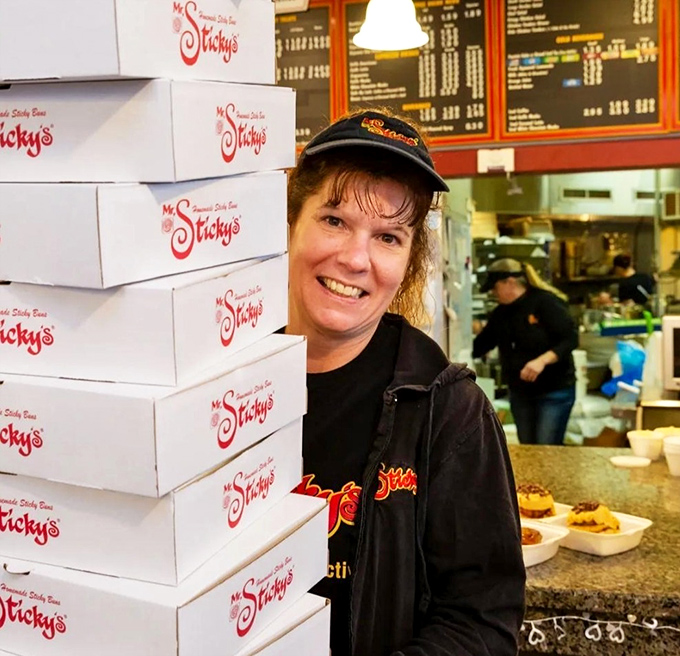 Stacks of to-go boxes ready for those wise enough to take extras home. The smile says it all—she's delivering happiness in cardboard form.