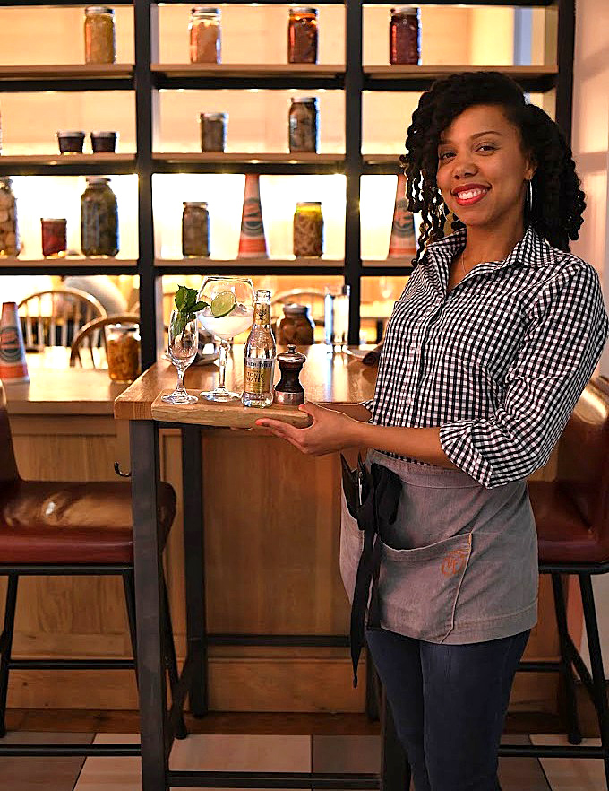 The display of preserved ingredients behind her isn't just decoration &ndash; it's a preview of the flavor complexity awaiting your palate.