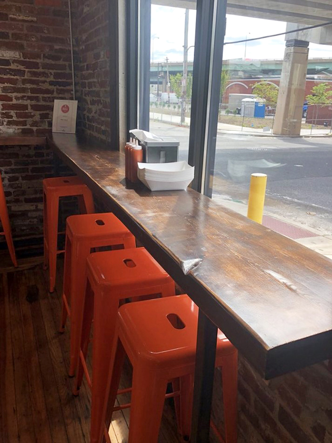 The window counter seating offers prime people-watching with those distinctive orange stools adding a pop of color against the rustic wood and brick.