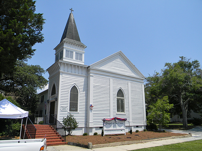 The Chapel of the Cross stands pristine against Carolina blue skies, its white clapboard exterior a testament to simpler times and enduring faith.
