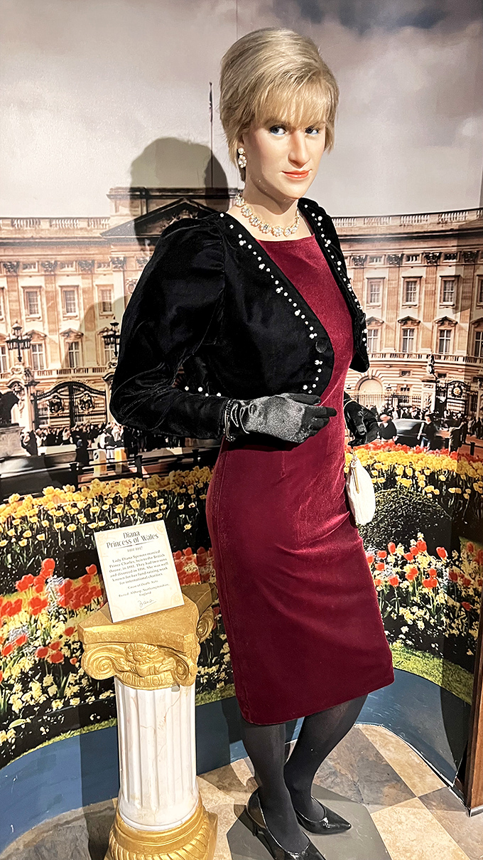 Princess Diana's wax figure captures her elegant style and compassionate gaze, standing before a backdrop of Buckingham Palace and vibrant gardens.