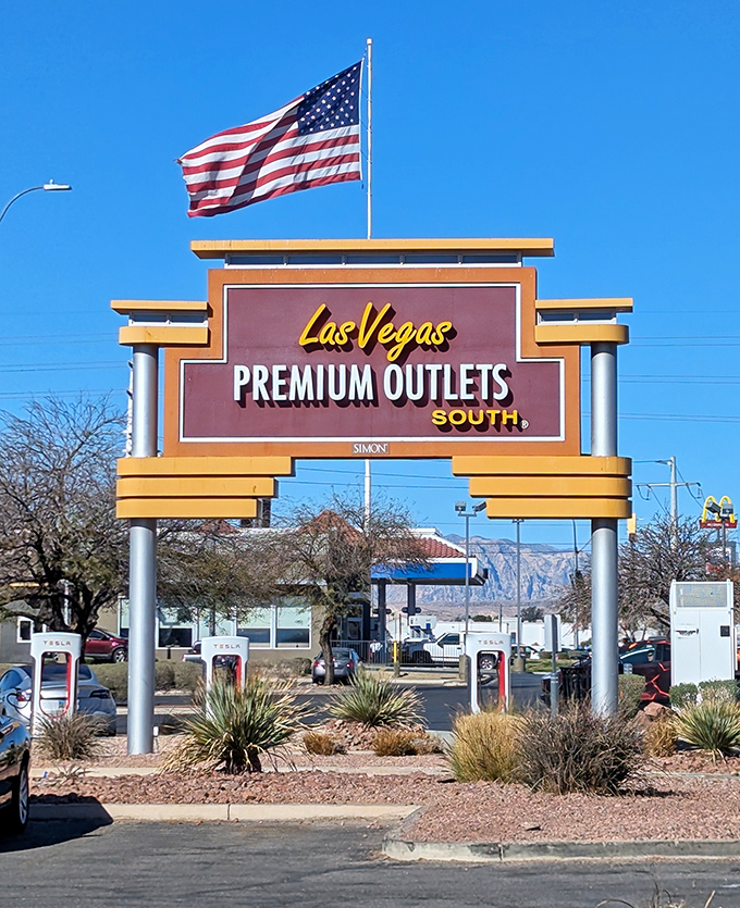 The iconic Las Vegas Premium Outlets South sign stands proudly against the desert sky, an American flag fluttering above like a beacon to bargain hunters.