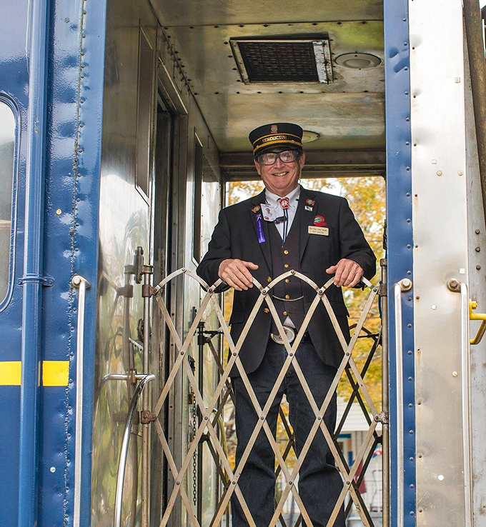 A uniformed conductor stands ready at his post, the human embodiment of railroad tradition and hospitality rolled into one smiling package.