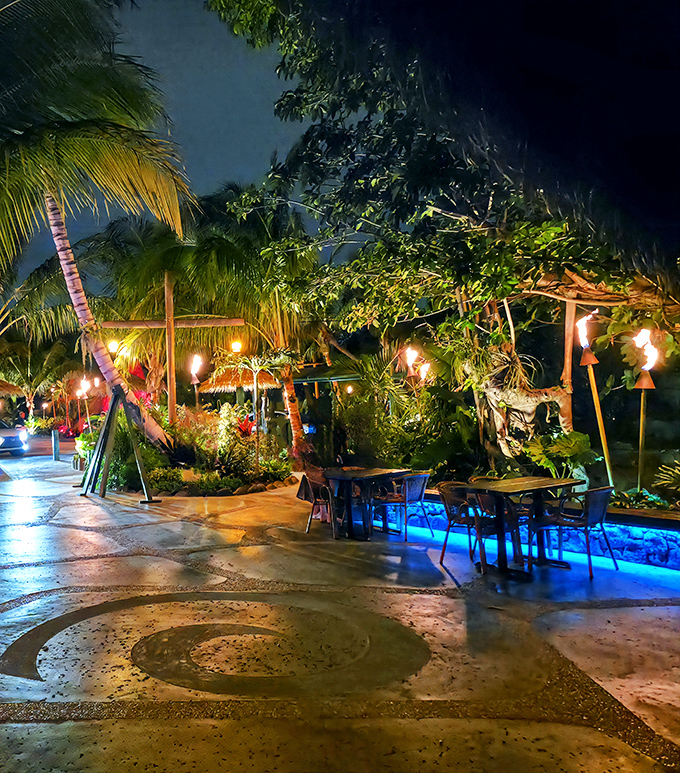 As night falls, the garden transforms into a blue-lit wonderland where tiki torches flicker against the Florida sky.