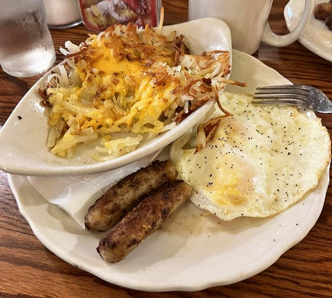 The perfect breakfast trinity &ndash; crispy hash browns crowned with melted cheese, sausage links, and an egg sunny-side up like morning on a plate.