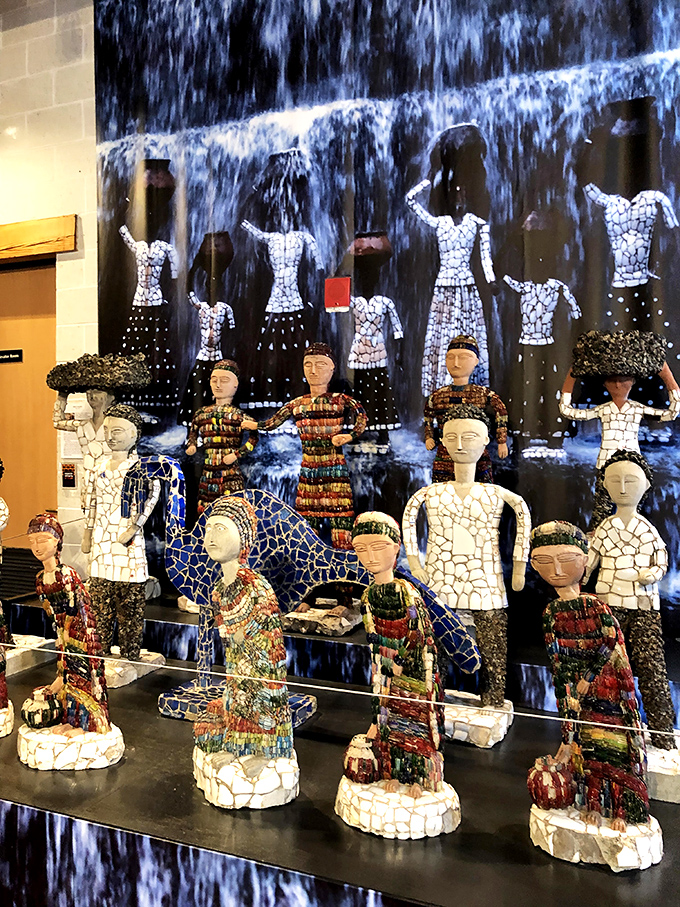 Mosaic figurines stand before a waterfall backdrop, their ceramic faces serene and knowing. Each unique figure tells its own story through carefully placed fragments and found objects.