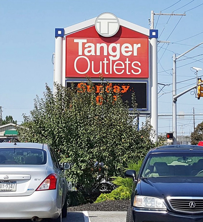 The red Tanger sign beckons from the highway – a retail lighthouse guiding bargain seekers safely to the shores of discounted designer goods.