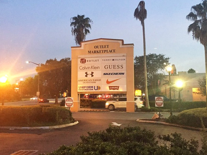 The entrance sign glows like a retail North Star at sunset, listing brand names that would normally send your credit card into hiding.
