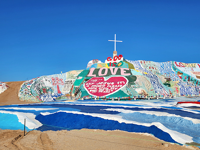 A vibrant mountain of paint and passion rises from the desert floor, topped with a cross and "LOVE" in bold letters.