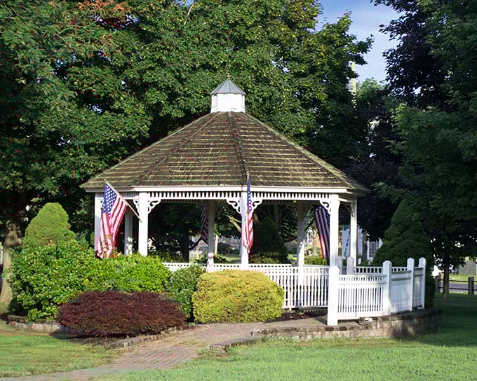 The town gazebo stands like a sentry of simpler times, draped in American flags and surrounded by the kind of greenery that makes you want to start a book club.