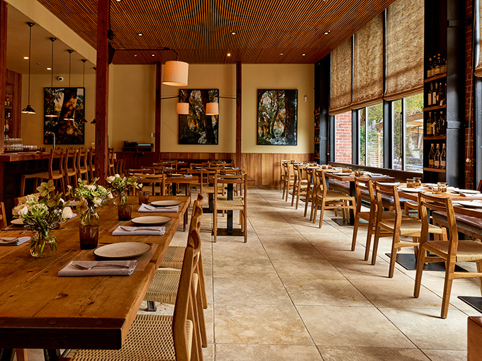 Sunlight streams through large windows, illuminating wooden tables and slatted ceilings in a dining room that feels both rustic and refined.