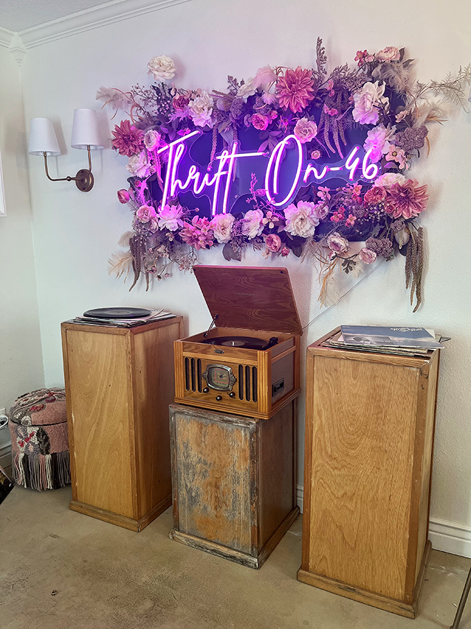 The neon "Thrift On 46" sign blooms amid artificial flowers, like a Vegas showgirl who decided to retire to a botanical garden.
