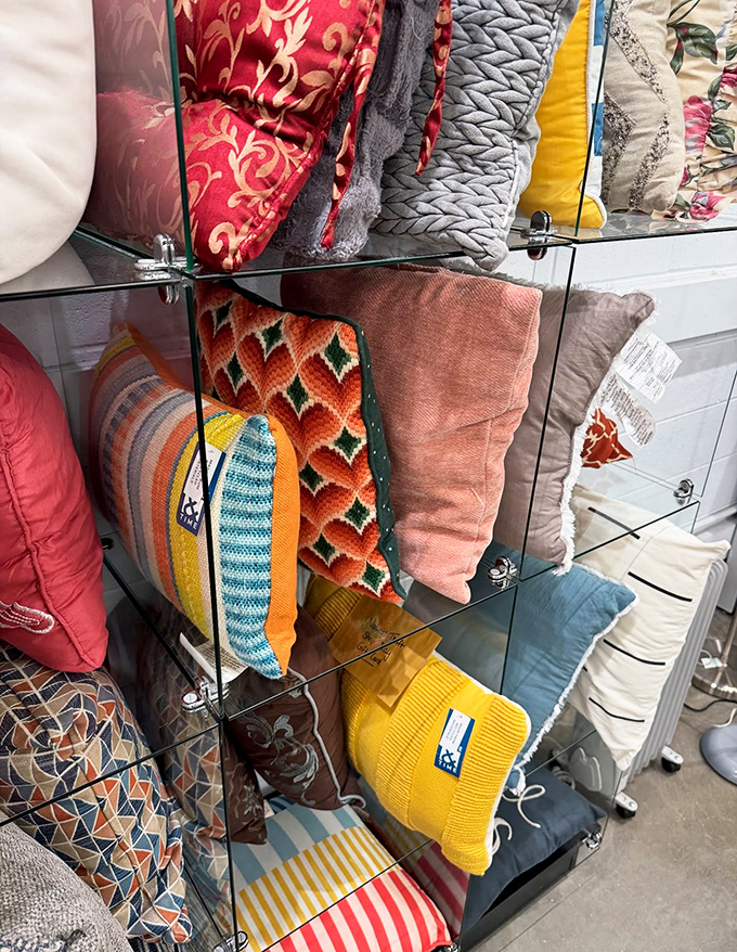 A kaleidoscope of decorative pillows stacked neatly behind glass &ndash; from vintage patterns to modern designs, each one waiting for its second chance to shine in someone's home.
