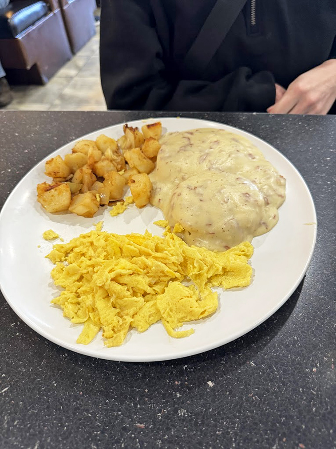 Behold the holy trinity of breakfast: golden home fries, fluffy scrambled eggs, and that legendary sausage gravy cascading over a hidden biscuit treasure.