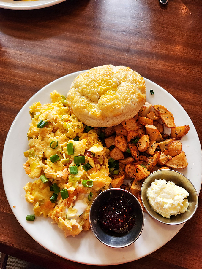 Breakfast perfection on a plate: scrambled eggs with green onions, seasoned potatoes, a fluffy biscuit, and sides of jam and butter. The morning ritual elevated.