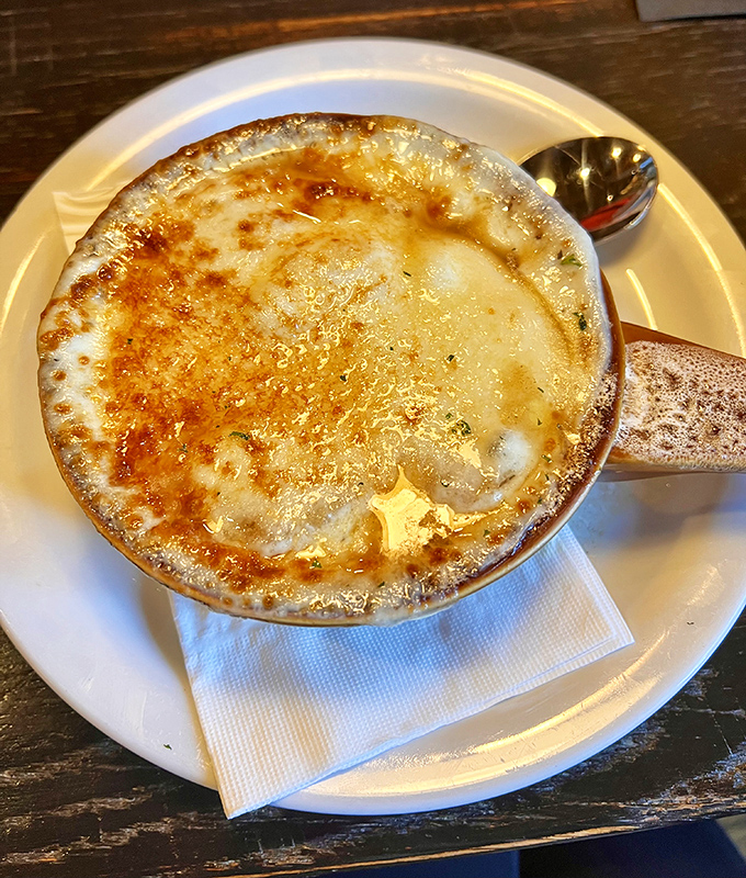 The legendary French onion soup arrives like a bubbling cauldron of comfort, its golden cheese canopy hiding a rich, savory treasure beneath.